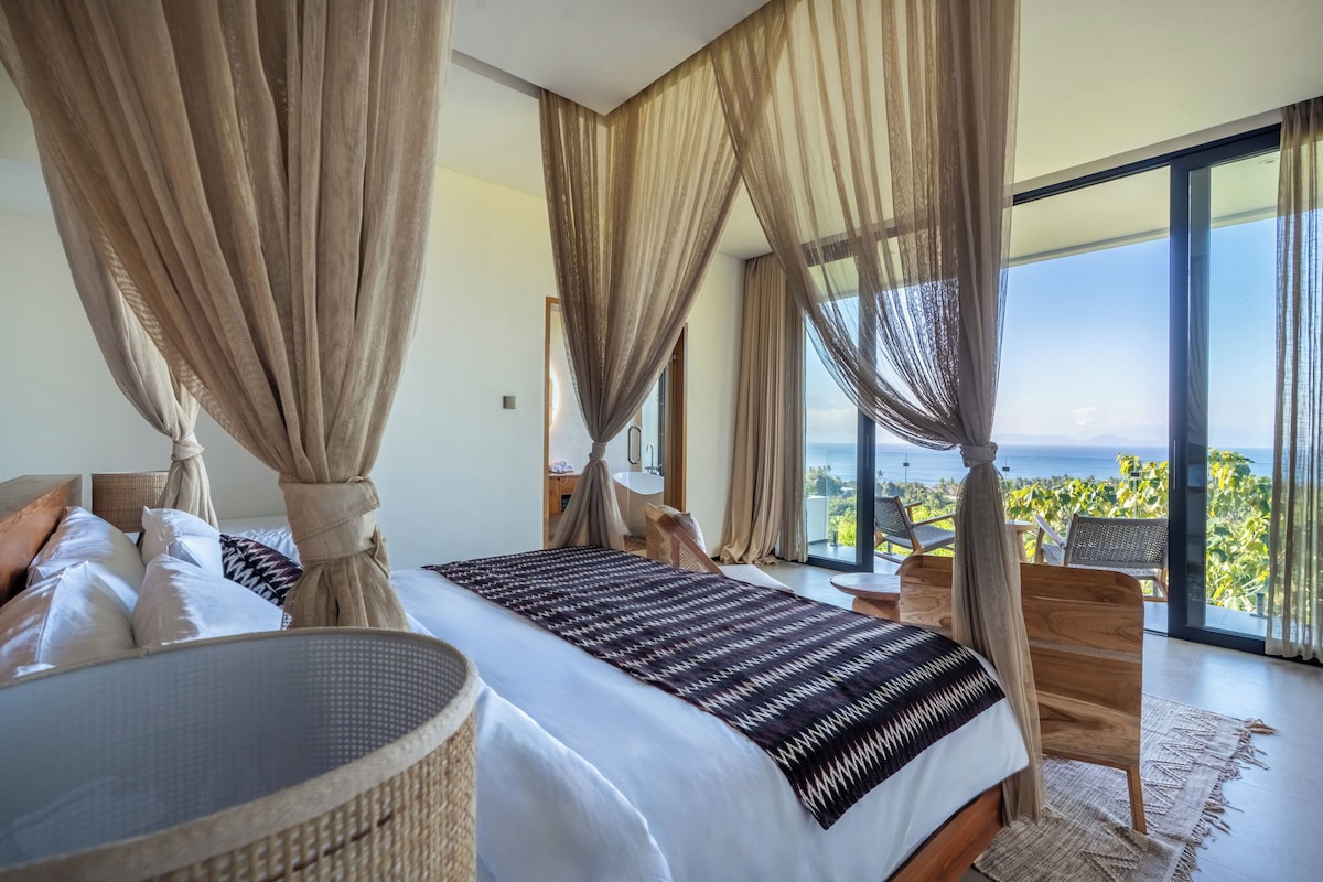 Quarto com uma cama grande, mesa de cabeceira, cadeira e vista para o oceano.