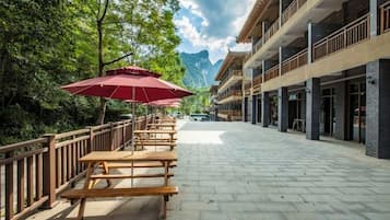 Uma passarela pavimentada com bancos de madeira e guarda-sóis vermelhos, ladeada por edifícios com varandas e lojas, com árvores e montanhas ao fundo.