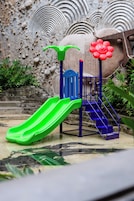Um parquinho com um escorrega verde, uma estrutura de escalada azul e uma decoração de flor vermelha.