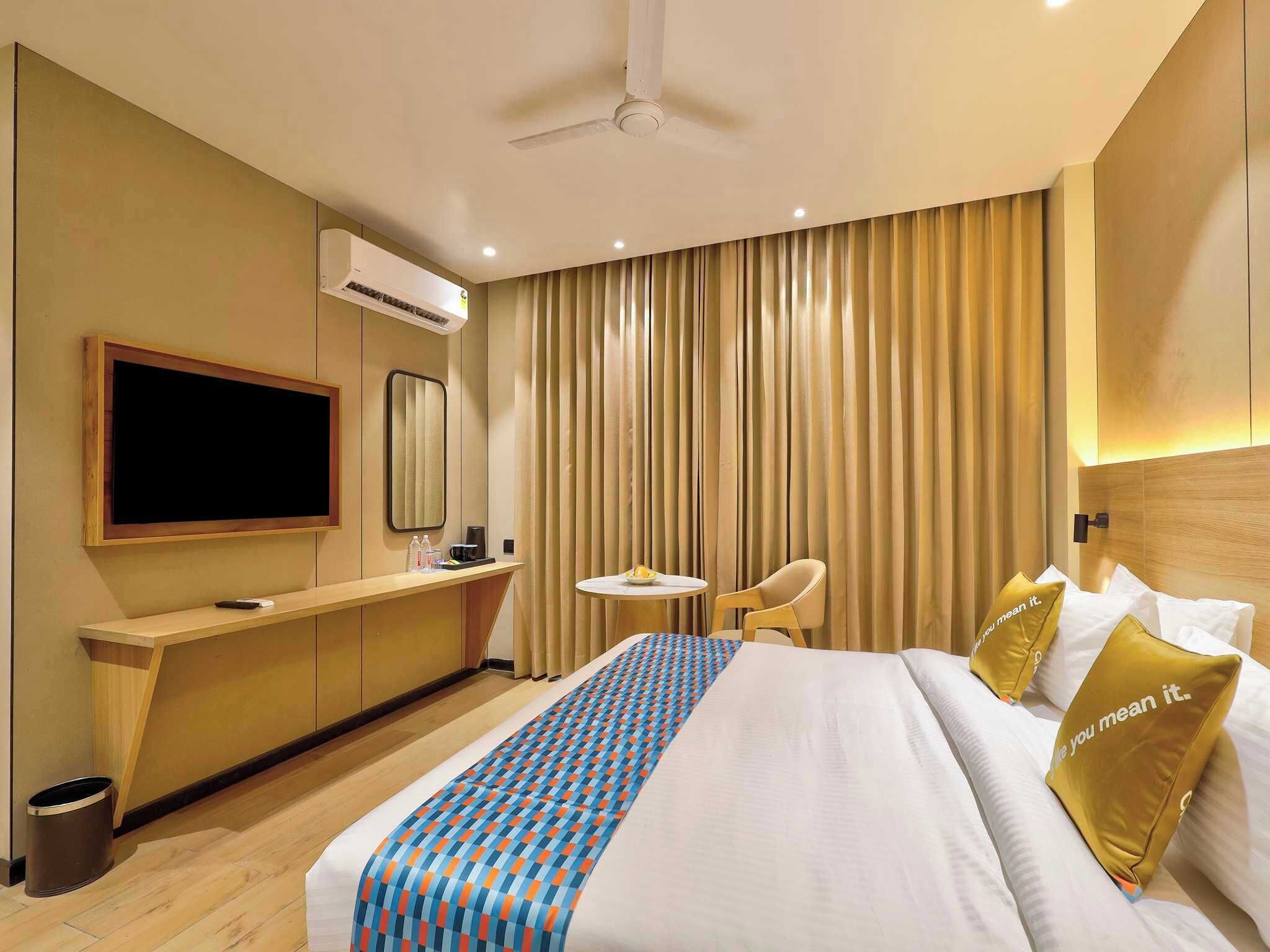 Quarto de hotel com cama, escrivaninha, cadeira, televisão e janela com cortinas.