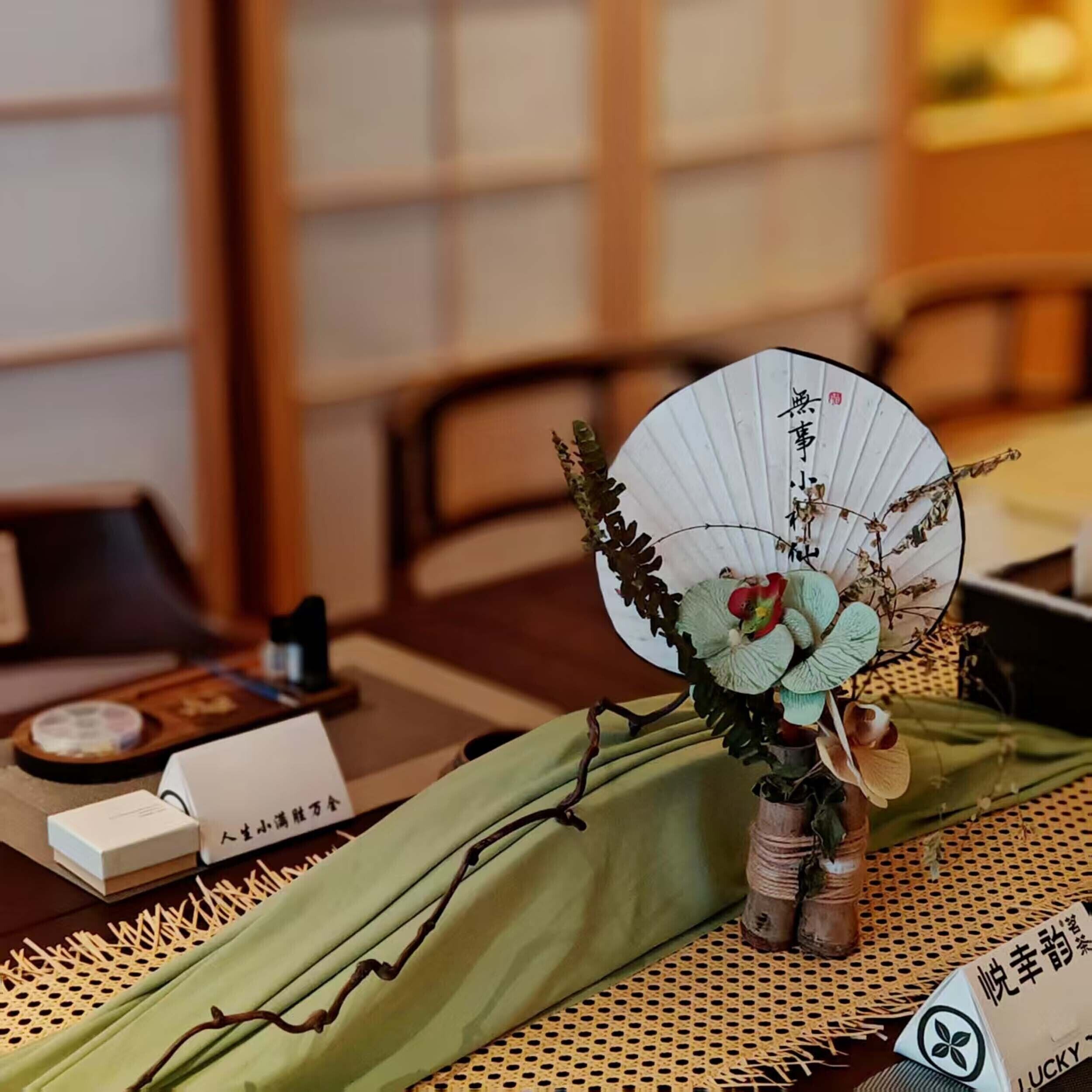 Um leque japonês tradicional com caligrafia, um pequeno vaso com flores e um esteira de bambu.