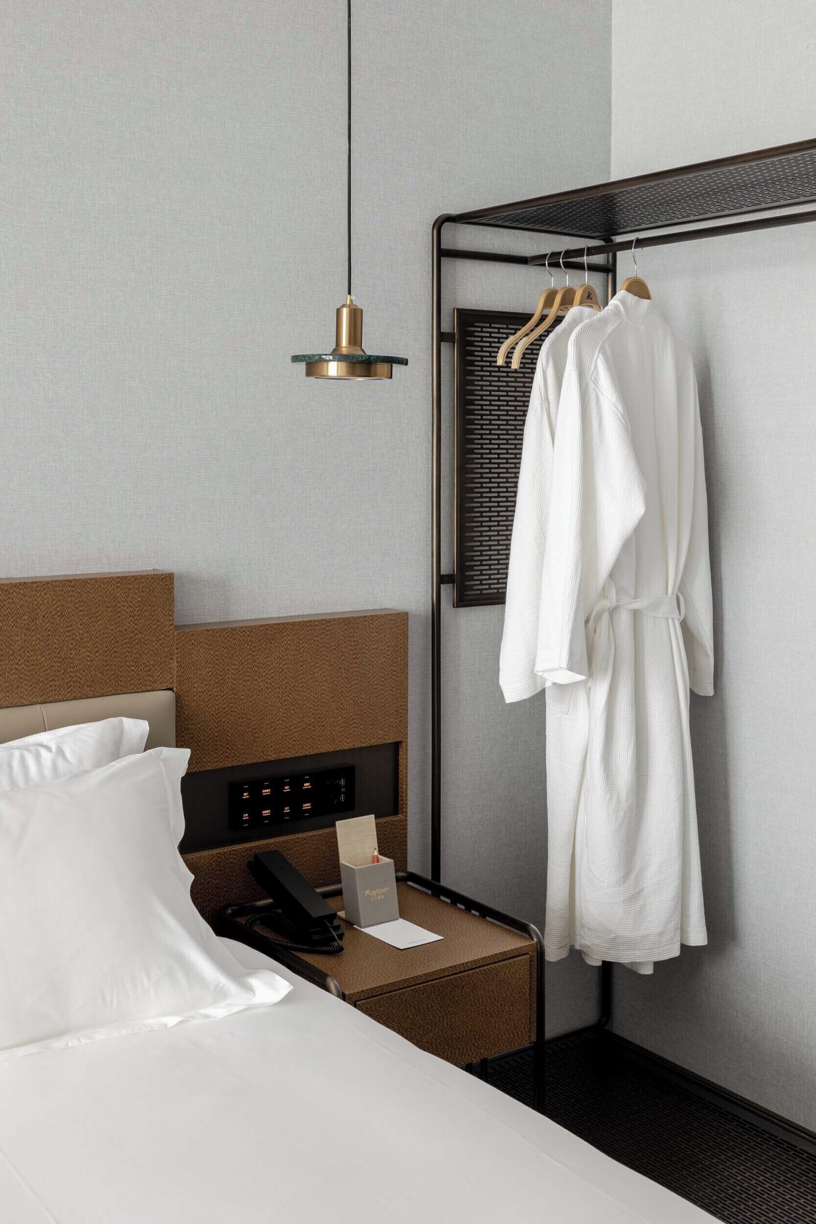 Quarto de hotel com cama, mesa de cabeceira com telefone e uma caixa, roupão pendurado em um suporte e uma luminária pendente.