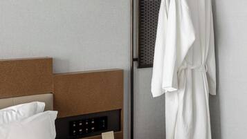 Quarto de hotel com cama, mesa de cabeceira com telefone e uma caixa, roupão pendurado em um suporte e uma luminária pendente.