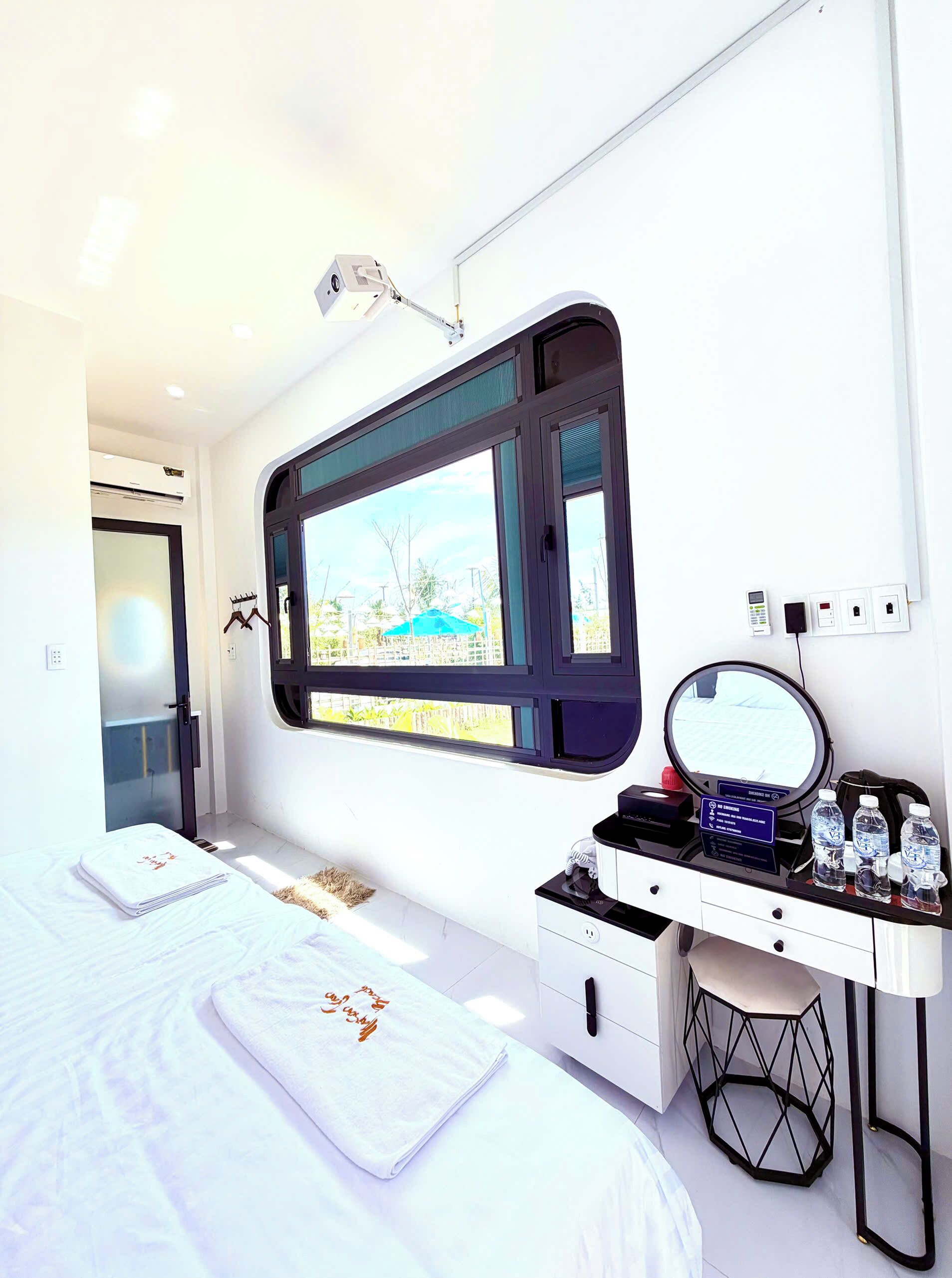 Quarto de hotel com uma grande janela, uma cama, uma mesa de cabeceira com espelho e um banquinho pequeno.