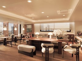 Uma área de refeições moderna com uma mesa de madeira, cadeiras e um banco. Há luminárias no teto e uma janela grande com vista.