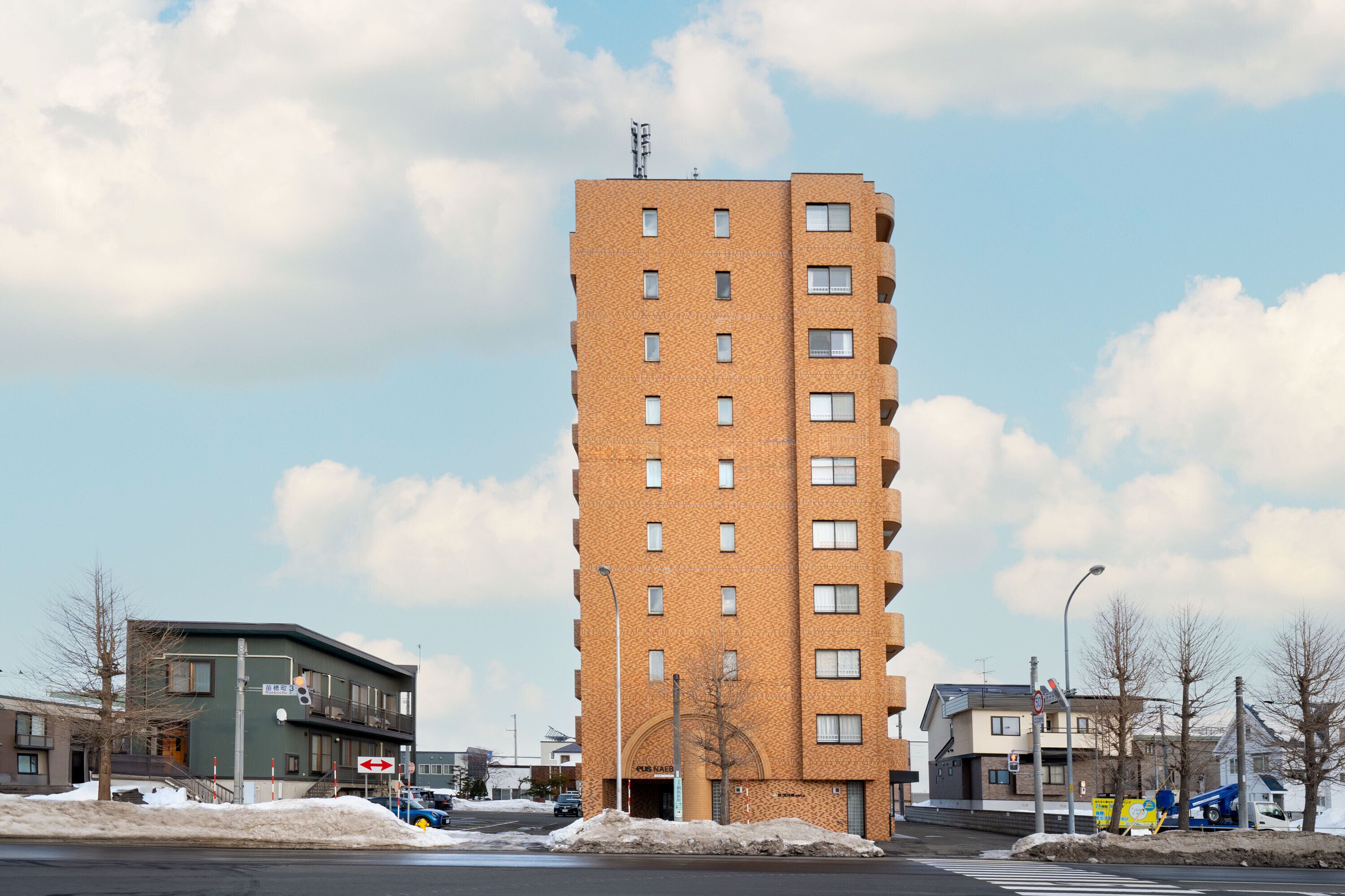 Um edifício alto de tijolos com vários andares e janelas, situado numa área urbana com neve, com outros edifícios e uma rua ao primeiro plano.