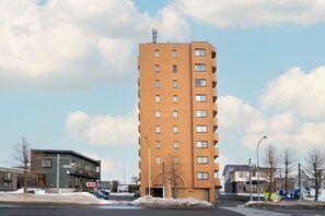 Um edifício alto de tijolos com vários andares e janelas, situado numa área urbana com neve, com outros edifícios e uma rua ao primeiro plano.