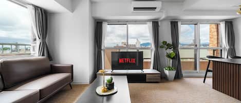 Uma sala de estar moderna com sofá, mesa de centro, TV exibindo 'NETFLIX' e uma varanda com vista.