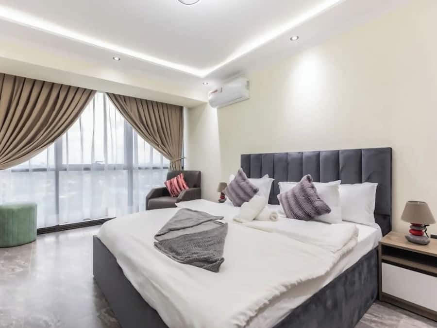 Quarto de hotel moderno com uma cama grande, uma poltrona, uma mesa de cabeceira e vista para a cidade.