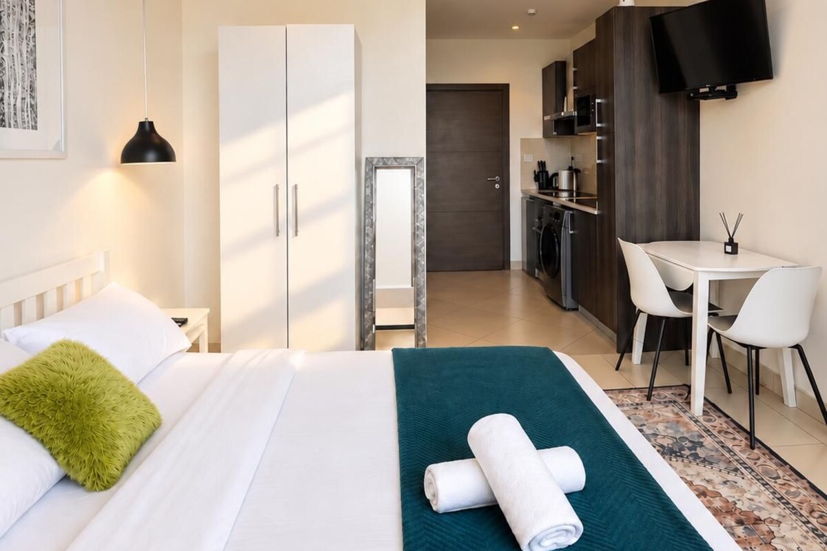 Quarto de hotel com cama, escrivaninha, cadeira, televisão e cozinha compacta.
