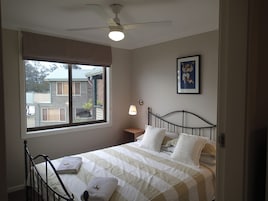 Quarto com cama, mesa de cabeceira, luminária na parede, ventilador de teto e janela com vista para uma casa.