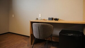 Um quarto pequeno e bem organizado, com uma escrivaninha de madeira, uma cadeira cinza, um cesto de lixo preto e uma tomada na parede.