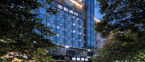 Um edifício moderno de hotel com fachada de vidro, sinalização iluminada e área externa bem cuidada com árvores e uma cerca.