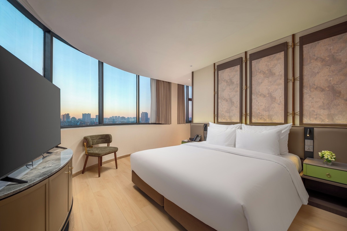 Quarto de hotel com uma cama grande, uma cadeira, uma escrivaninha com televisão de tela plana e vista para a cidade através de grandes janelas.