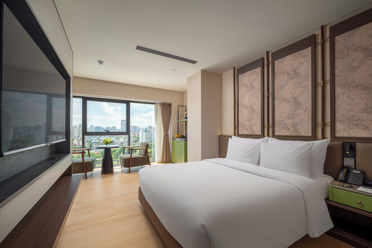 Quarto de hotel com cama grande, escrivaninha, cadeira e vista para a cidade, além de uma janela ampla.