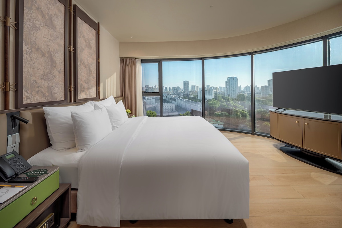 Quarto de hotel com uma cama grande, televisão de tela plana, escrivaninha com telefone e vista para a cidade através de grandes janelas.