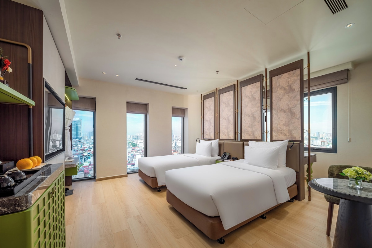 Quarto de hotel com duas camas, uma mesa pequena, vista para a cidade e uma cozinha compacta.