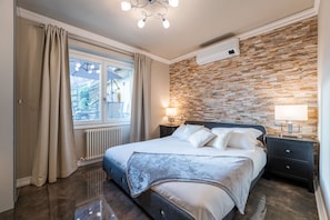 Quarto com uma cama grande, parede de pedra, janela com cortinas e mesa de cabeceira com abajur.