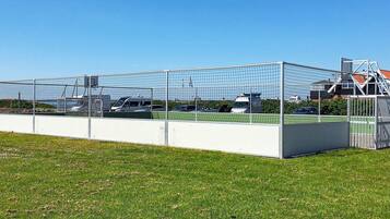 Um campo esportivo cercado, com um edifício branco e uma construção de telhado vermelho ao fundo.