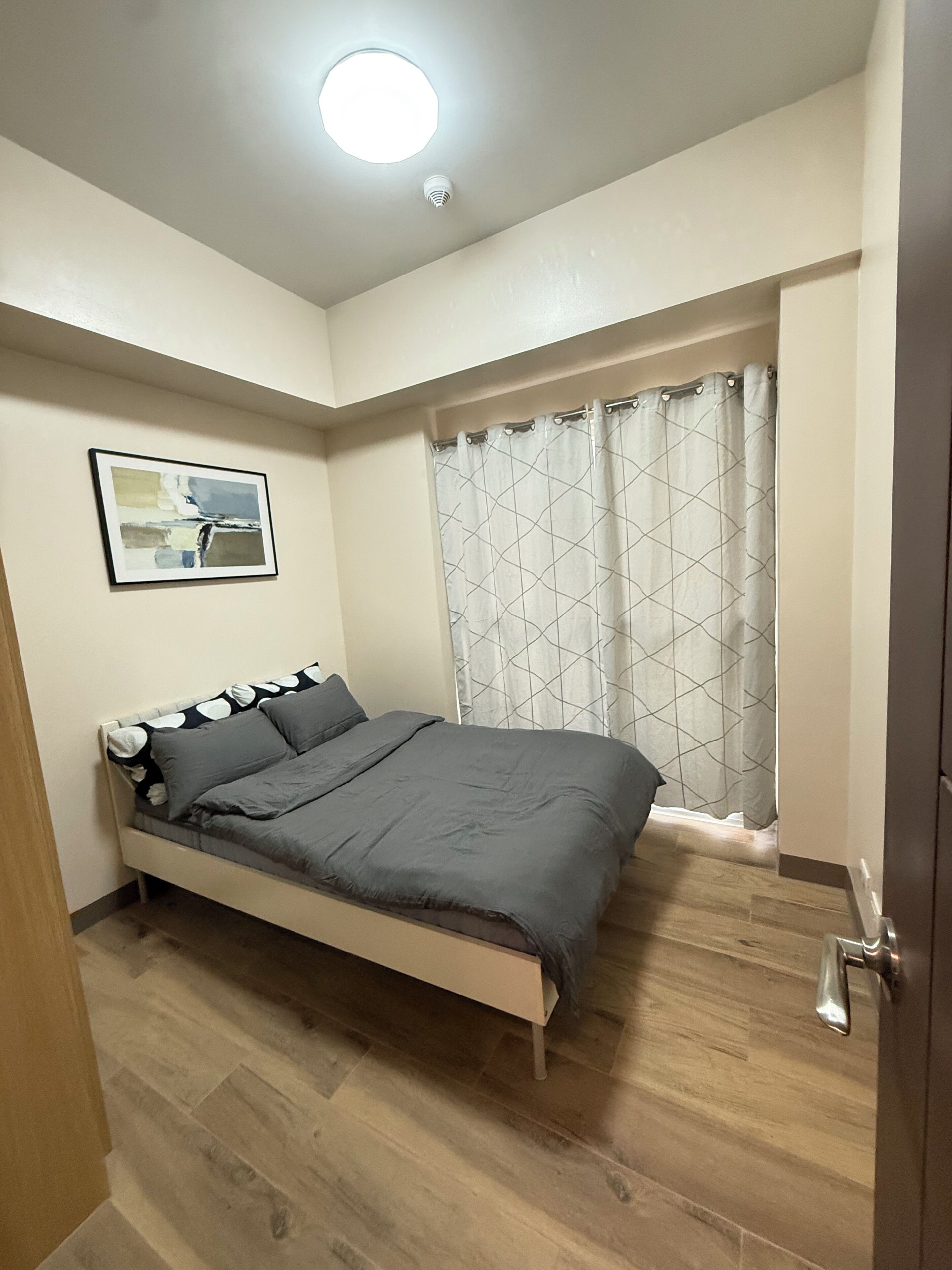 Quarto com cama, um quadro na parede, iluminação no teto e uma janela com cortina.