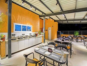 Uma área de refeições ao ar livre com mesas, cadeiras, um balcão de buffet e vista para o céu noturno.