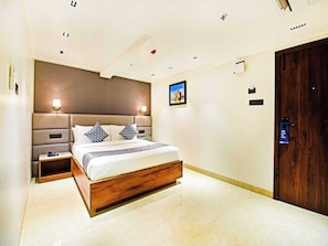 Quarto de hotel moderno com uma cama grande, mesas de cabeceira, um criado-mudo, cabeceira de madeira, um quadro na parede e uma porta com puxador.