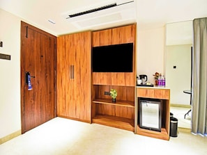 Quarto de hotel com guarda-roupa de madeira, televisão de tela plana, pequeno frigorífico e máquina de café.