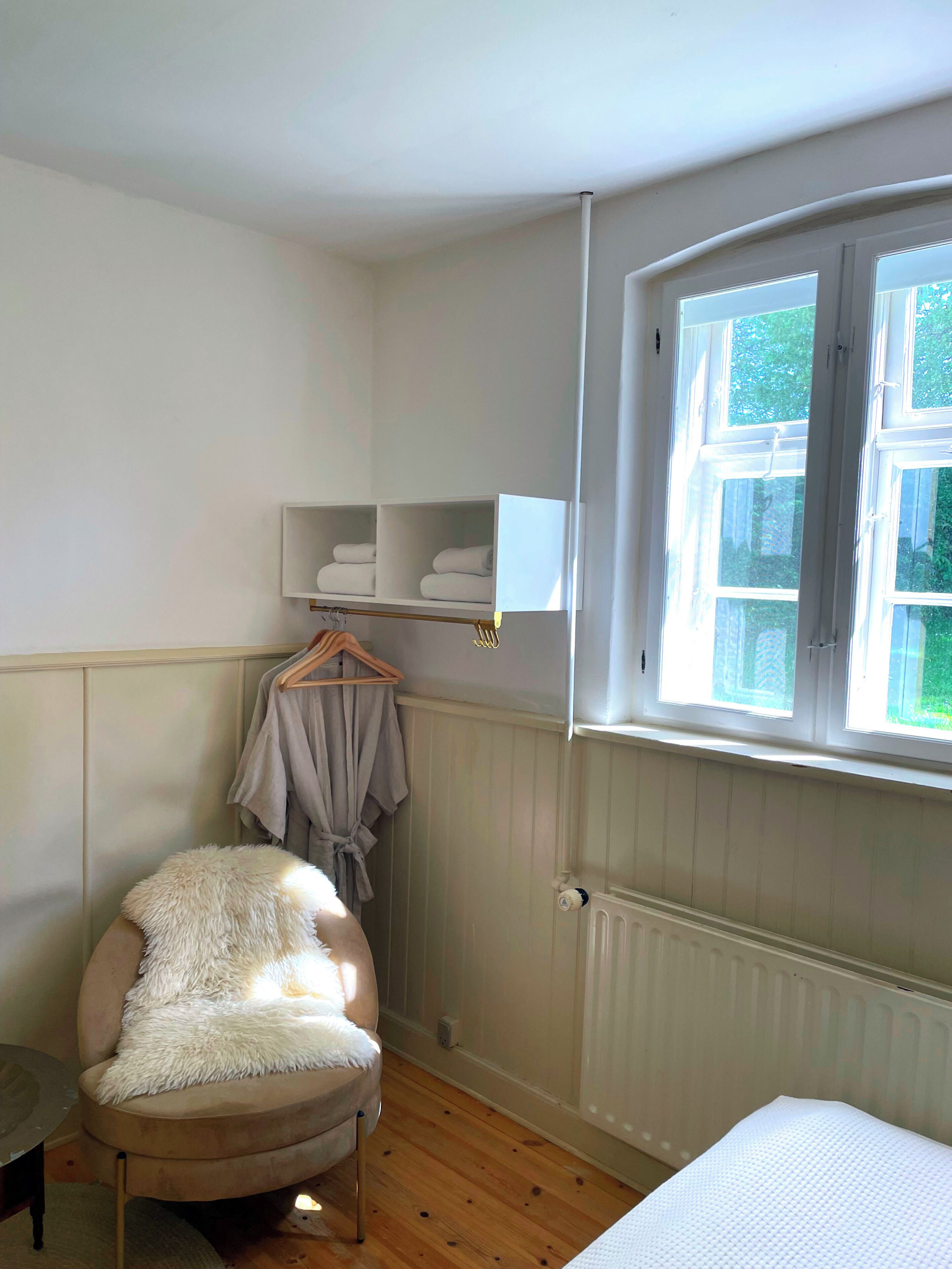 Quarto com cama, uma cadeira com travesseiro fofinho, radiador, janela com cortinas e prateleira com toalhas dobradas.