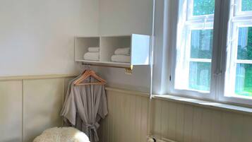 Quarto com cama, uma cadeira com travesseiro fofinho, radiador, janela com cortinas e prateleira com toalhas dobradas.