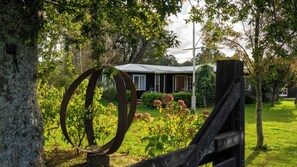 Uma casa rústica com uma escultura de metal em primeiro plano, cercada por vegetação exuberante e um gramado bem cuidado.