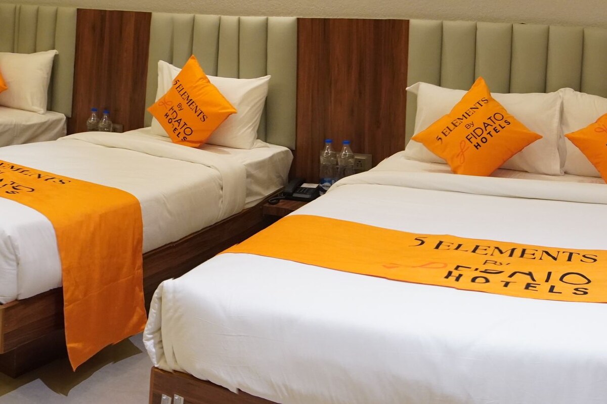 Quarto de hotel com duas camas, cada uma com colcha branca e detalhes em laranja, incluindo almofadas e um cobertor.