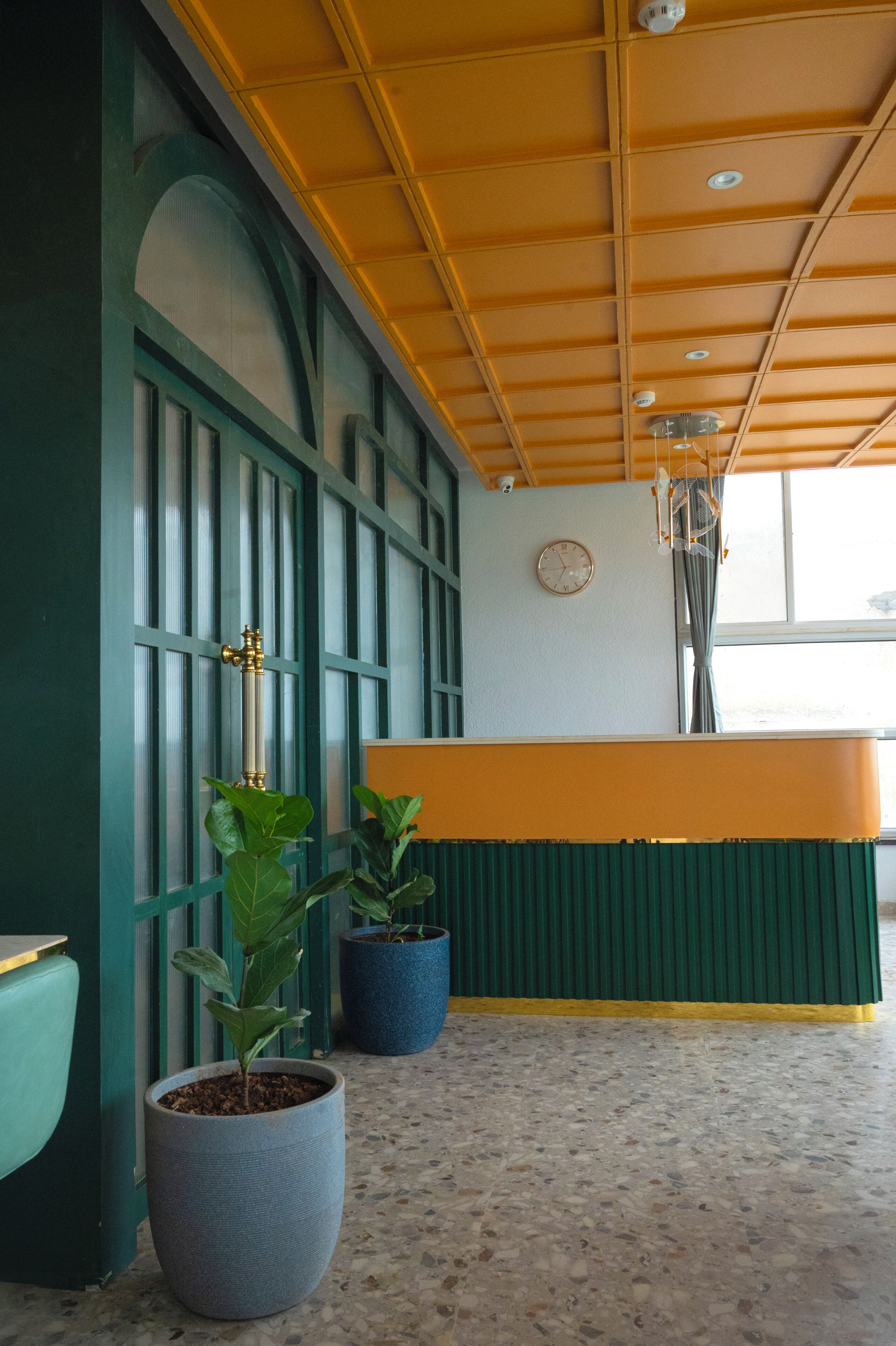 Uma área de recepção com esquema de cores verde e amarelo, um relógio na parede e plantas em vasos.