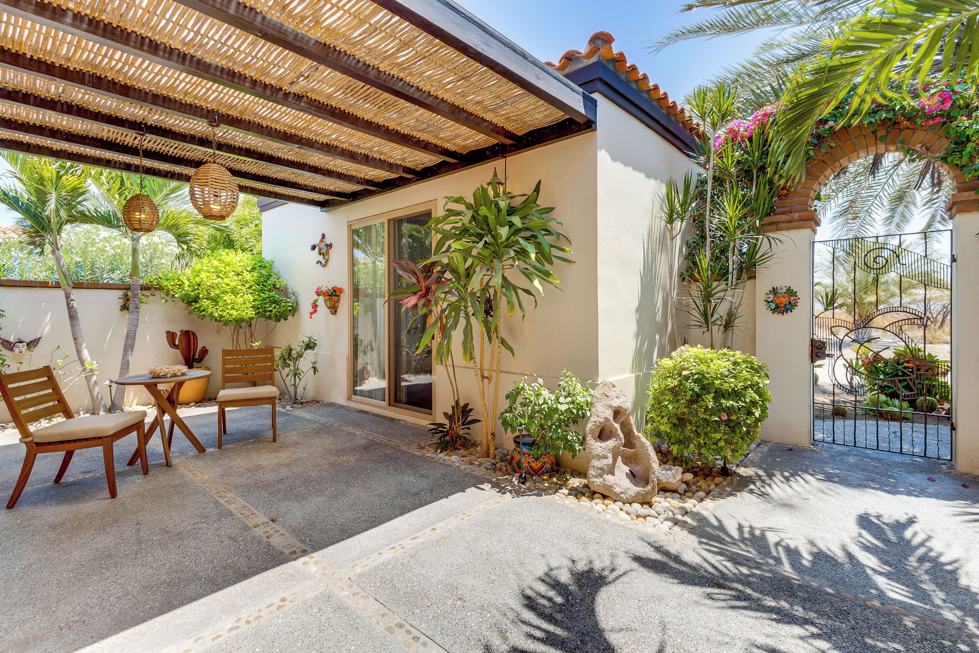 Uma área de varanda coberta com mesa e cadeiras, cercada por vasos de plantas e um muro de pedra decorativo.