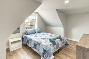 Quarto com teto inclinado, uma cama com colcha, mesa de cabeceira e janela com vista para casas.