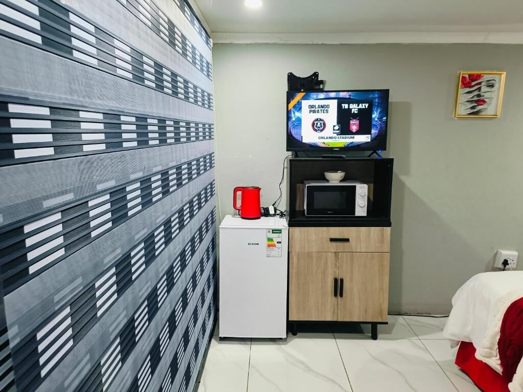Quarto de hotel com televisão mostrando uma partida esportiva, minibar e cama com cobertor vermelho.