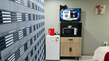 Quarto de hotel com televisão mostrando uma partida esportiva, minibar e cama com cobertor vermelho.