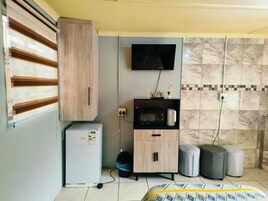 Quarto compacto com cama, pequena área de cozinha, televisão fixada na parede e revestimento de azulejos.