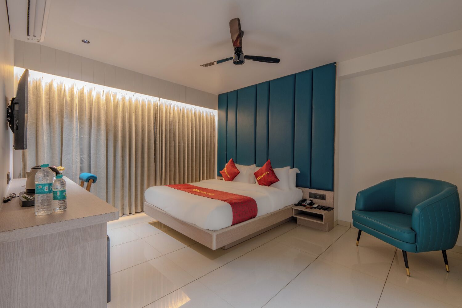 A modern hotel room with a bed, a desk with water bottles, a blue armchair, and a ceiling fan.