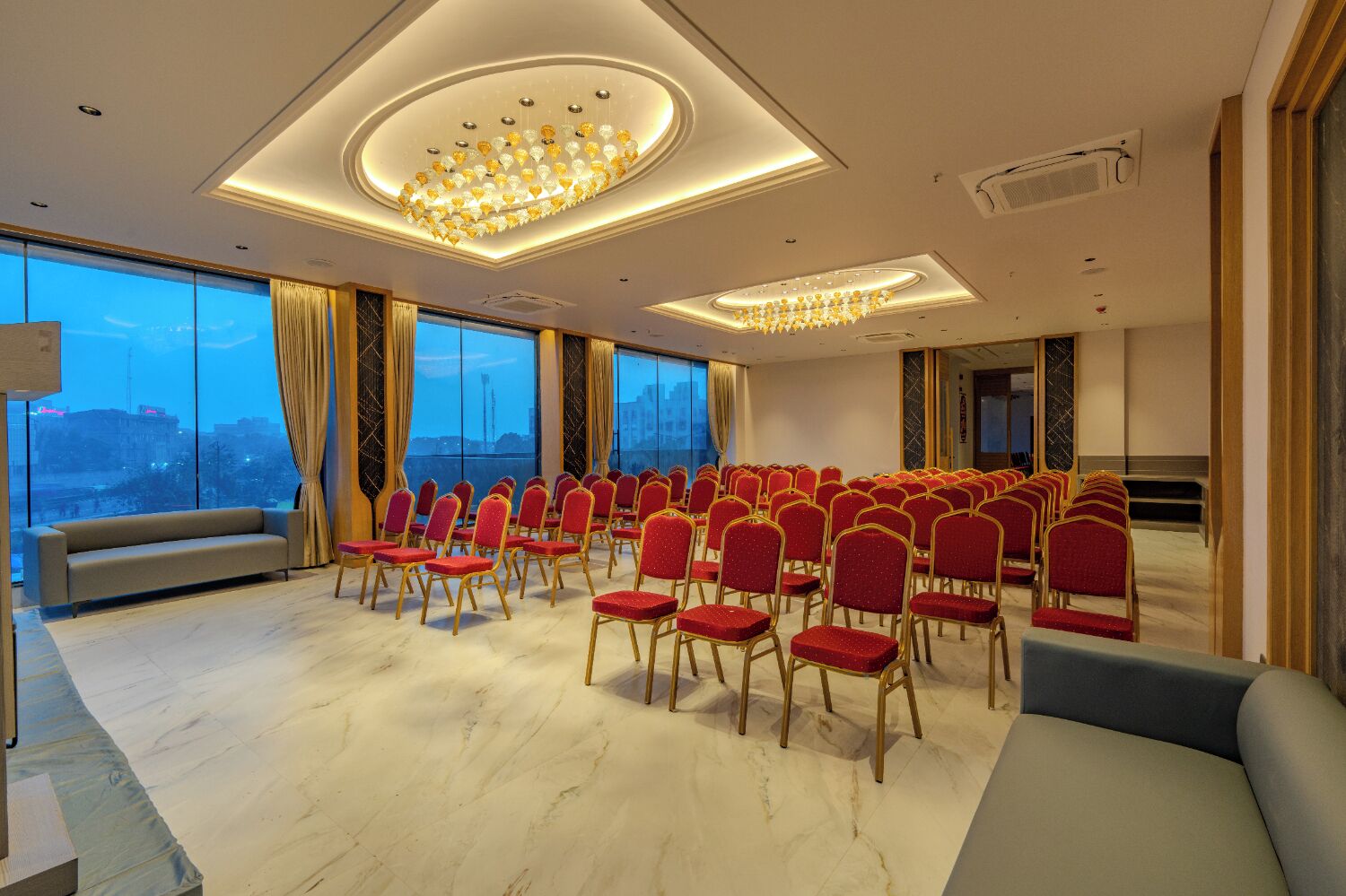A conference room with rows of red chairs, a large chandelier, and a city view through large windows.