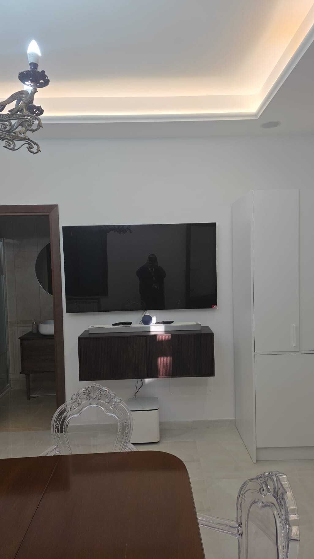 Uma sala de estar moderna com uma televisão de tela grande fixada na parede, uma mesa de jantar com cadeiras e um lustre.