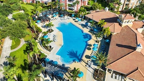 Vista aérea de um resort com uma piscina central, várias áreas de estar com guarda-sóis e diversos edifícios com telhados de telha vermelha.