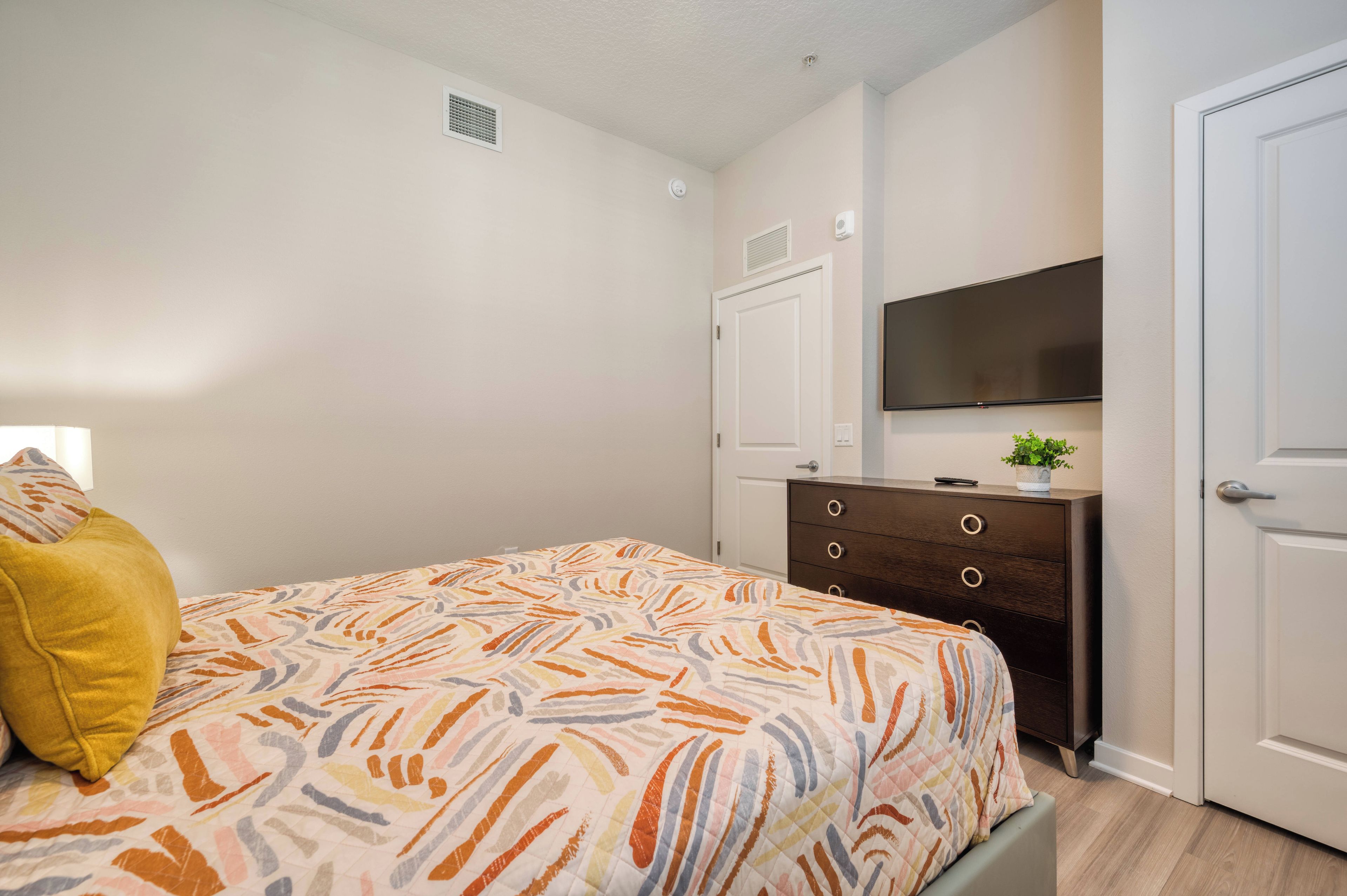 Quarto com cama, penteadeira, televisão fixada na parede e uma porta.