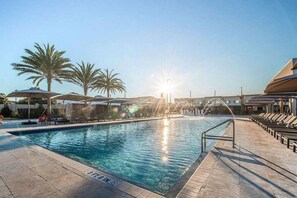 Piscina com espreguiçadeiras e guarda-sóis, cercada por palmeiras.