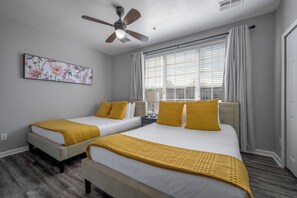 Quarto de hotel com duas camas, ventilador no teto, janela com cortinas e uma obra de arte floral na parede.
