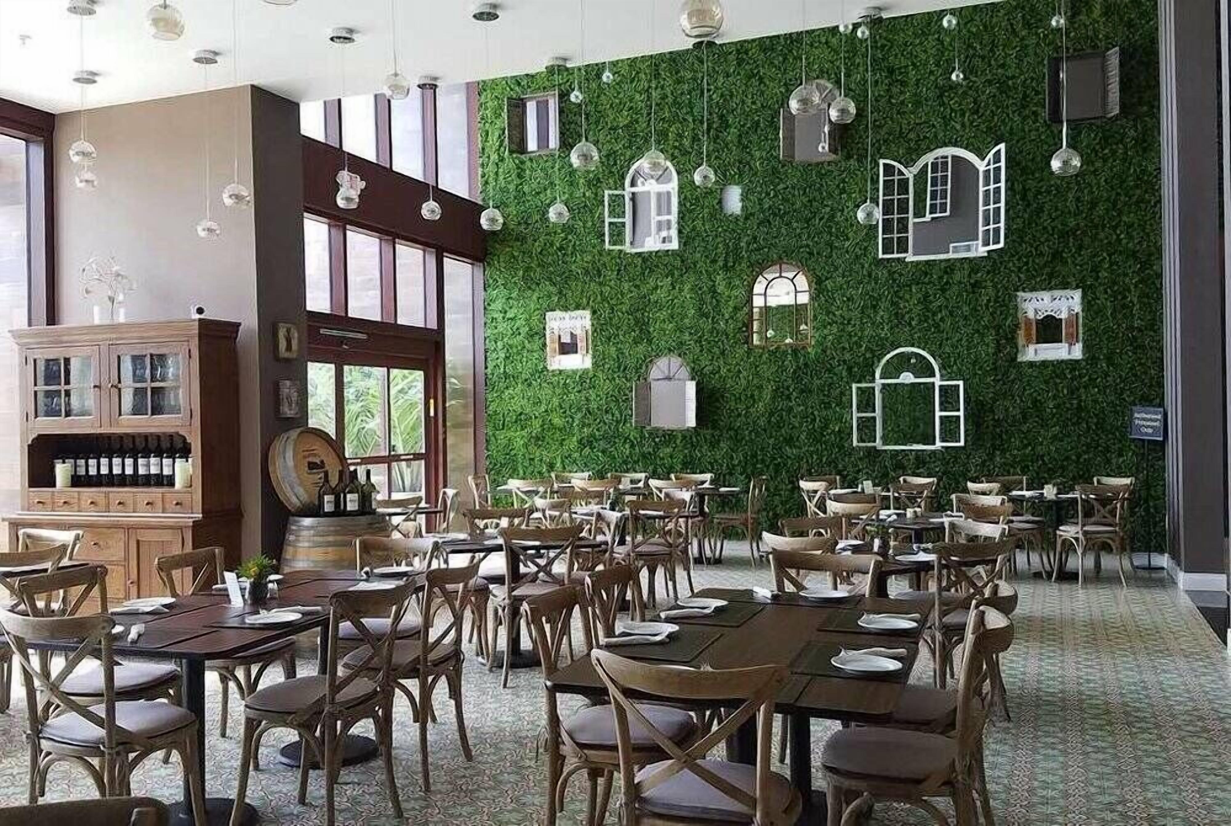 Restaurante com várias mesas e cadeiras, um armário de madeira com garrafas de vinho e uma parede verde com janelas decorativas.