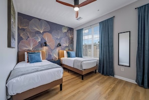 Quarto de hotel com duas camas, ventilador de teto, janela grande com cortinas e um mural na parede.