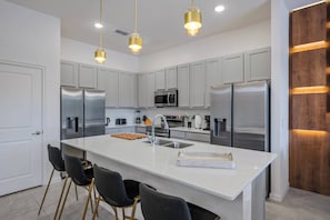 Uma cozinha moderna com armários brancos, eletrodomésticos de aço inoxidável, uma ilha central com banquetas e iluminação pendente.
