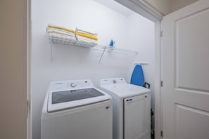 Uma área de lavanderia com duas máquinas de lavar brancas, um ferro de passar azul e uma prateleira com toalhas dobradas.