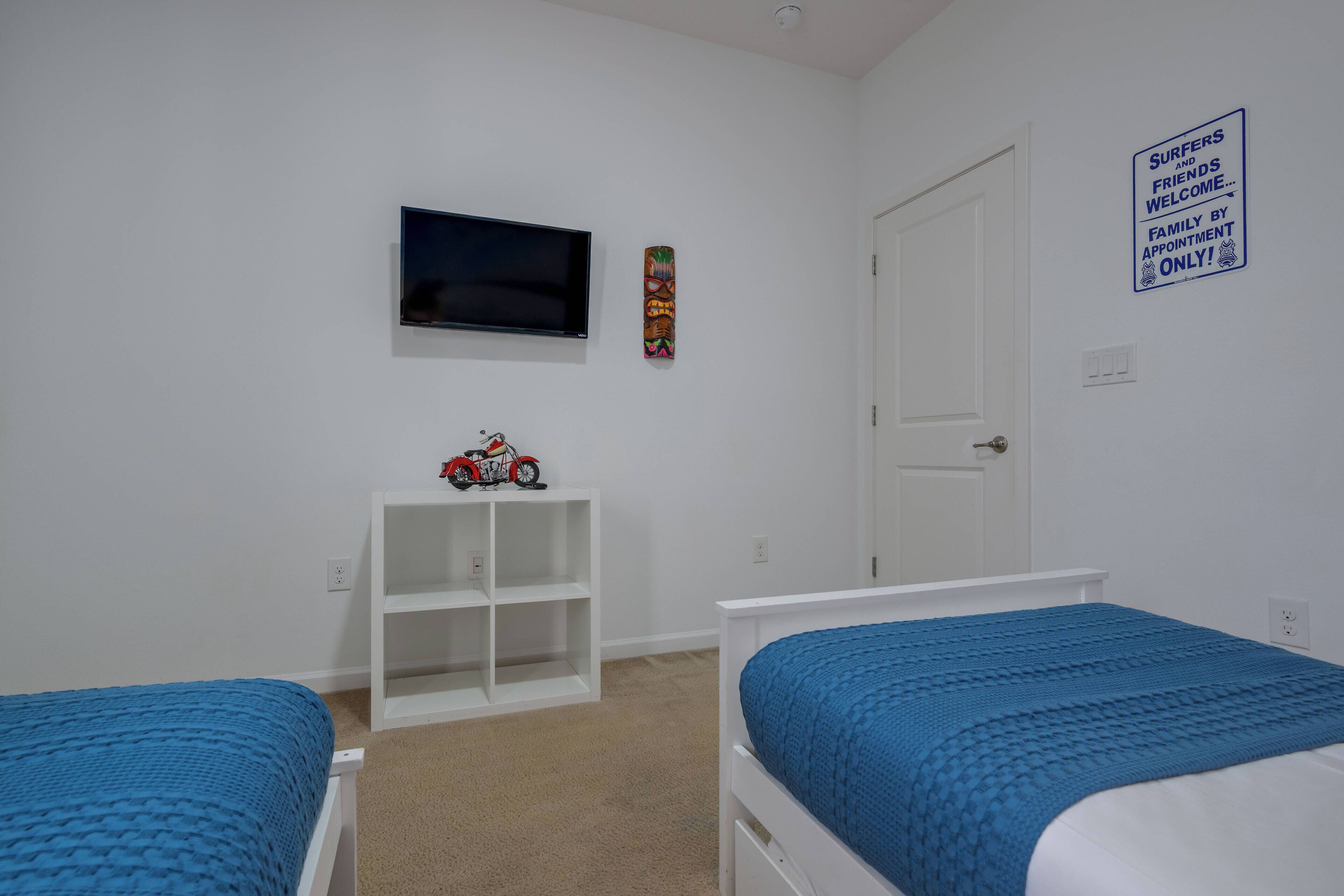 Quarto com duas camas, uma televisão, uma estante de brinquedos e uma porta.