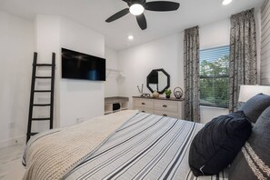 Um quarto com uma cama, televisão, penteadeira, espelho, janela com cortinas e ventilador de teto.